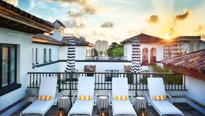 Terraço no telhado com cadeiras de lounge e guarda-sóis listrados, com vista para a paisagem urbana ao pôr do sol.
