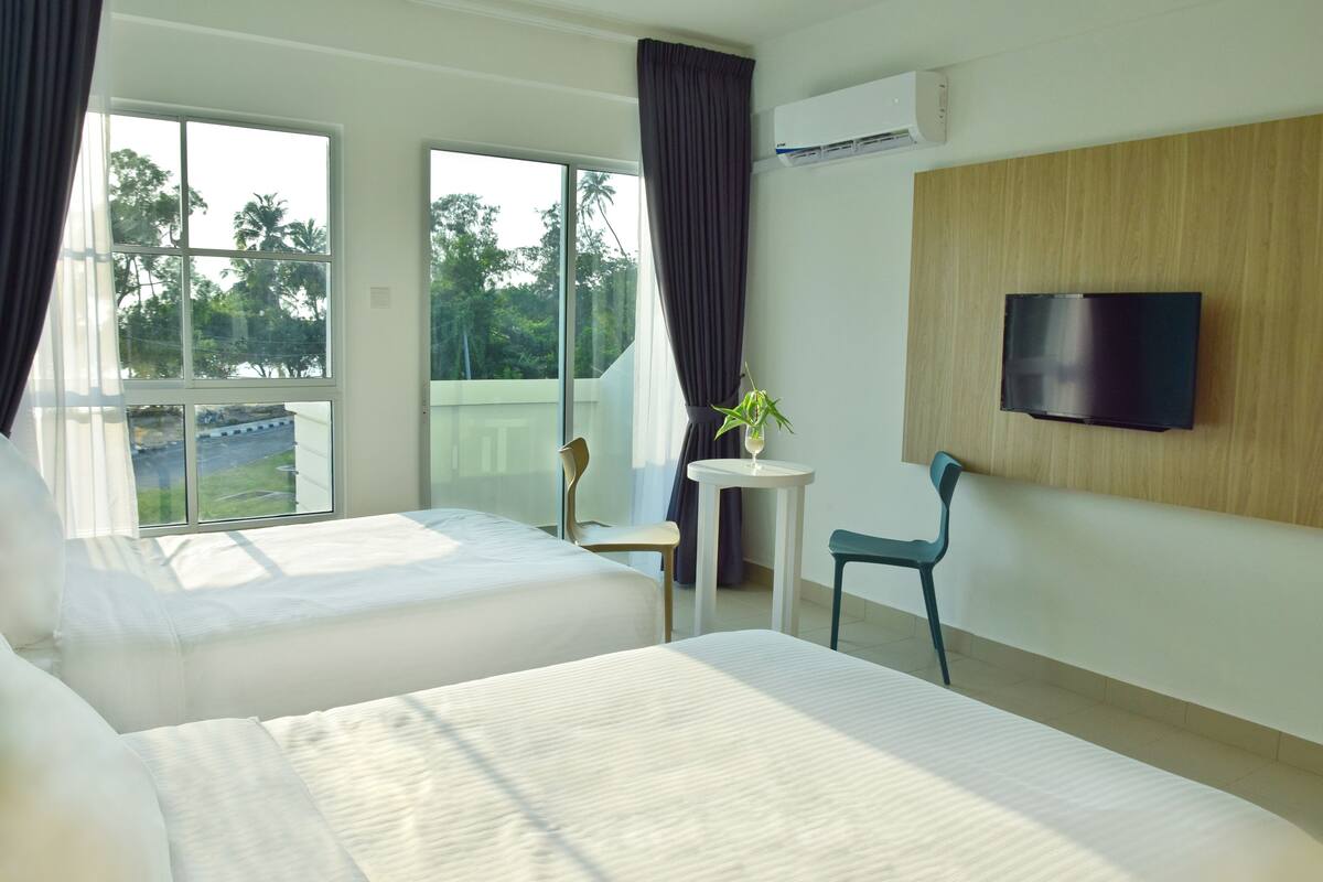 Quarto de hotel com duas camas, uma mesinha com uma planta, uma televisão na parede e uma janela grande com cortinas.