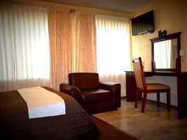 Quarto de hotel com uma cama, uma poltrona de couro marrom, uma escrivaninha de madeira com cadeira e uma televisão fixada na parede.