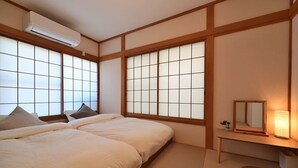 Um quarto tradicional japonês com tatames, uma cama com roupa de cama branca, uma mesa de cabeceira com espelho e uma luminária, e uma pequena planta em vaso.