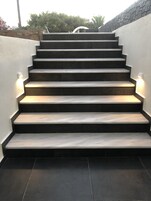 Escadas modernas iluminadas com parede de pedra e vegetação ao fundo.