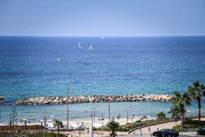 Vista costeira com um molhe rochoso, praia e veleiros na água.