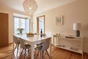 Uma sala de jantar com uma mesa branca, cadeiras cinzentas, uma luminária trançada e um aparador com abajur.