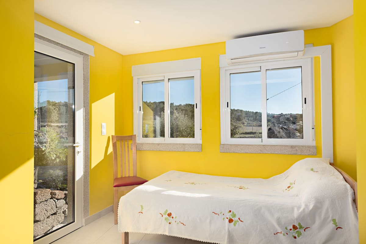 Quarto com paredes amarelas, uma cama com colcha floral, duas janelas com molduras brancas e uma cadeira.