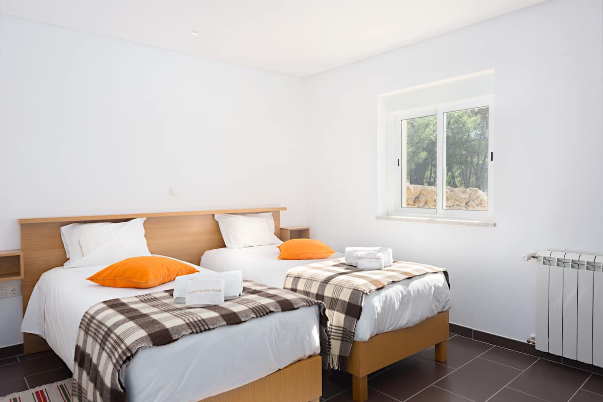 Quarto com duas camas, roupa de cama branca, almofadas laranja e uma janela com vista para árvores.