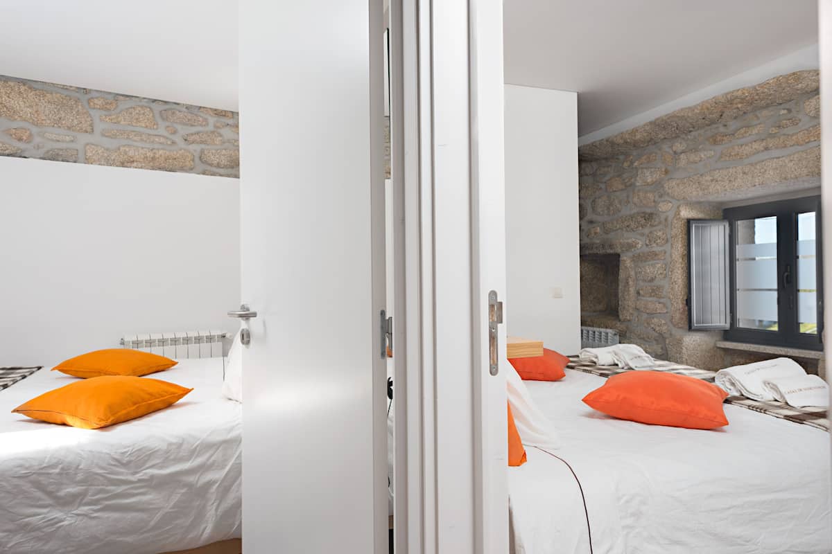 Quarto de hotel com duas camas, roupa de cama branca, almofadas laranja, parede de pedra e uma janela.