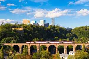 Um trem atravessa uma ponte de pedra com várias arcadas, cercado por vegetação e edifícios urbanos ao fundo.