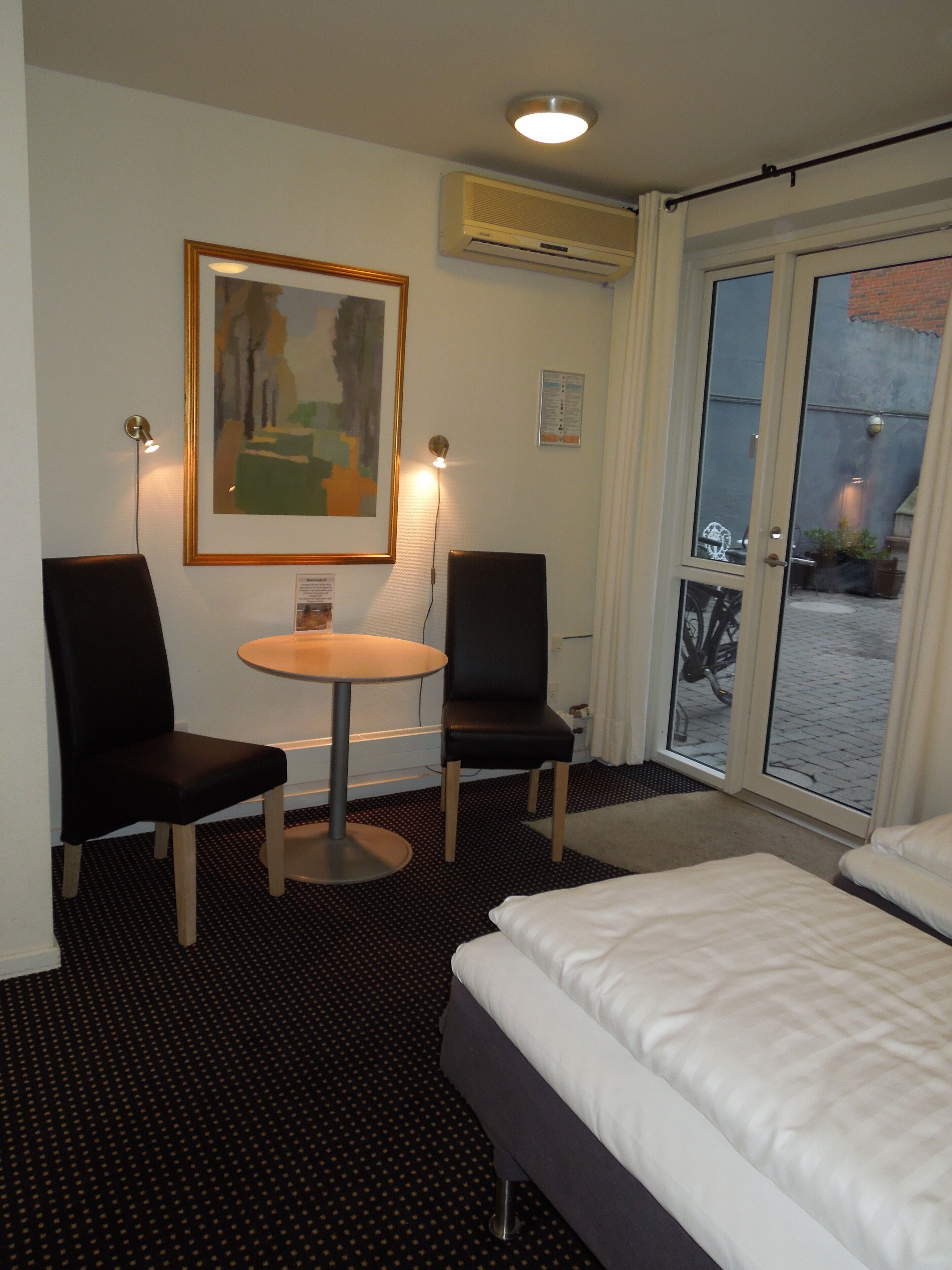 Quarto de hotel com uma cama, duas cadeiras, uma mesa pequena e um quadro na parede.