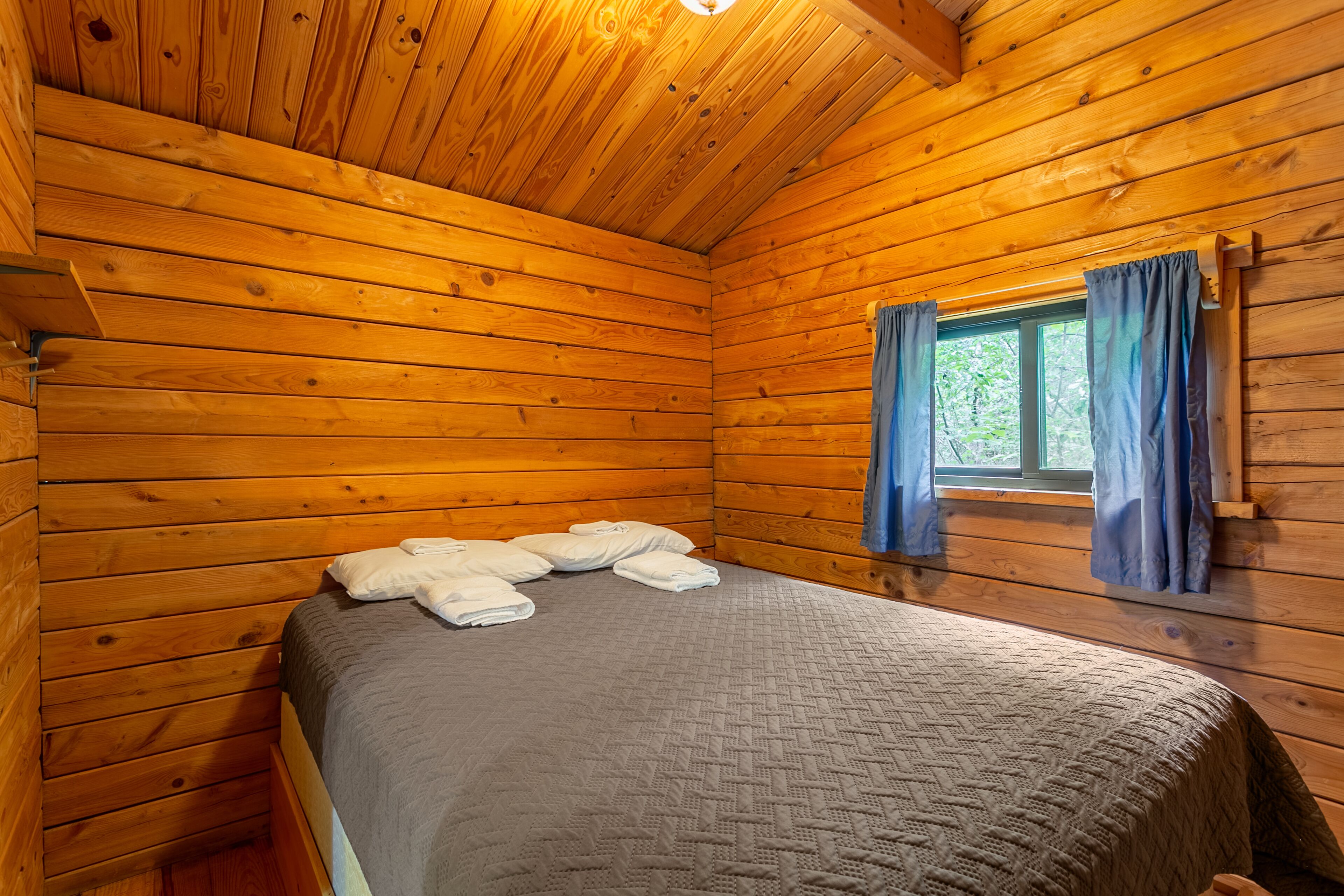 Quarto de cabana de madeira com uma cama, uma janela com cortinas e teto de madeira.