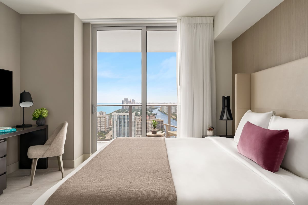 Quarto de hotel moderno com uma cama grande, uma mesa e uma cadeira. O quarto tem vista para um horizonte urbano e um corpo d'água.