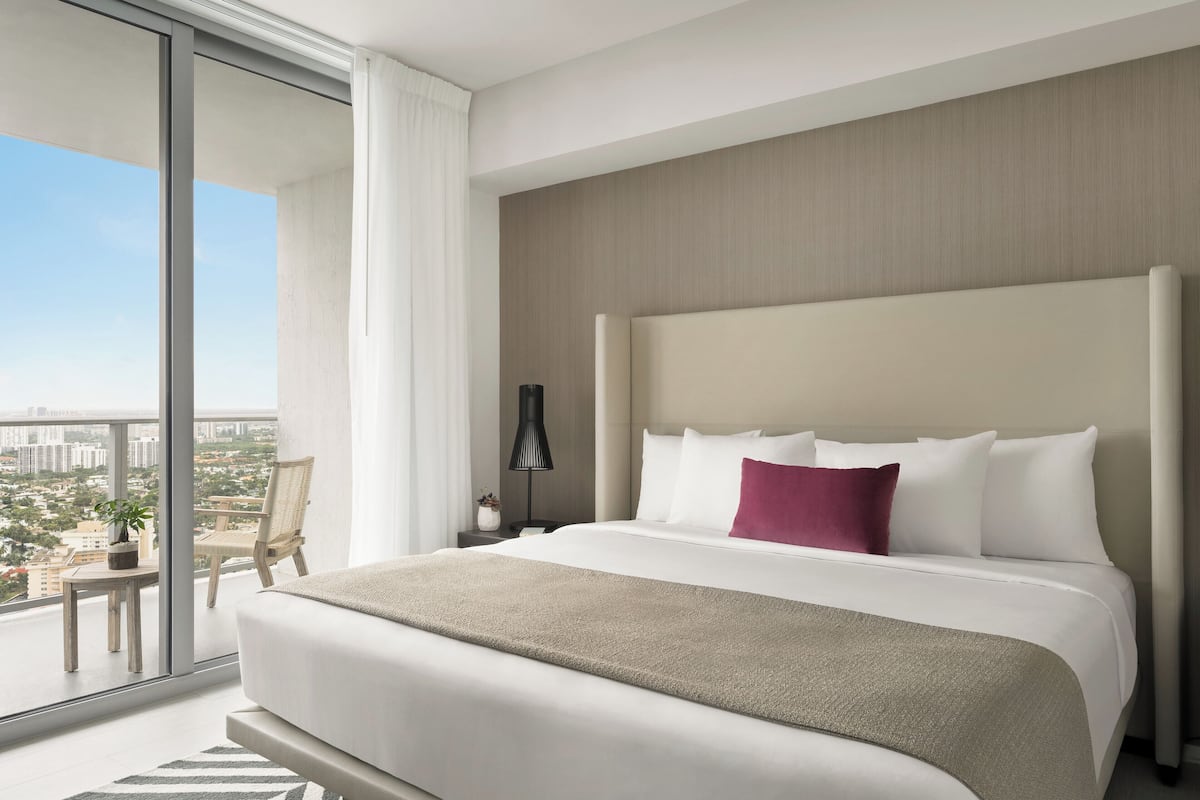 Quarto de hotel moderno com uma cama grande, vista da cidade, varanda com uma cadeira e mesa de cabeceira com abajur.