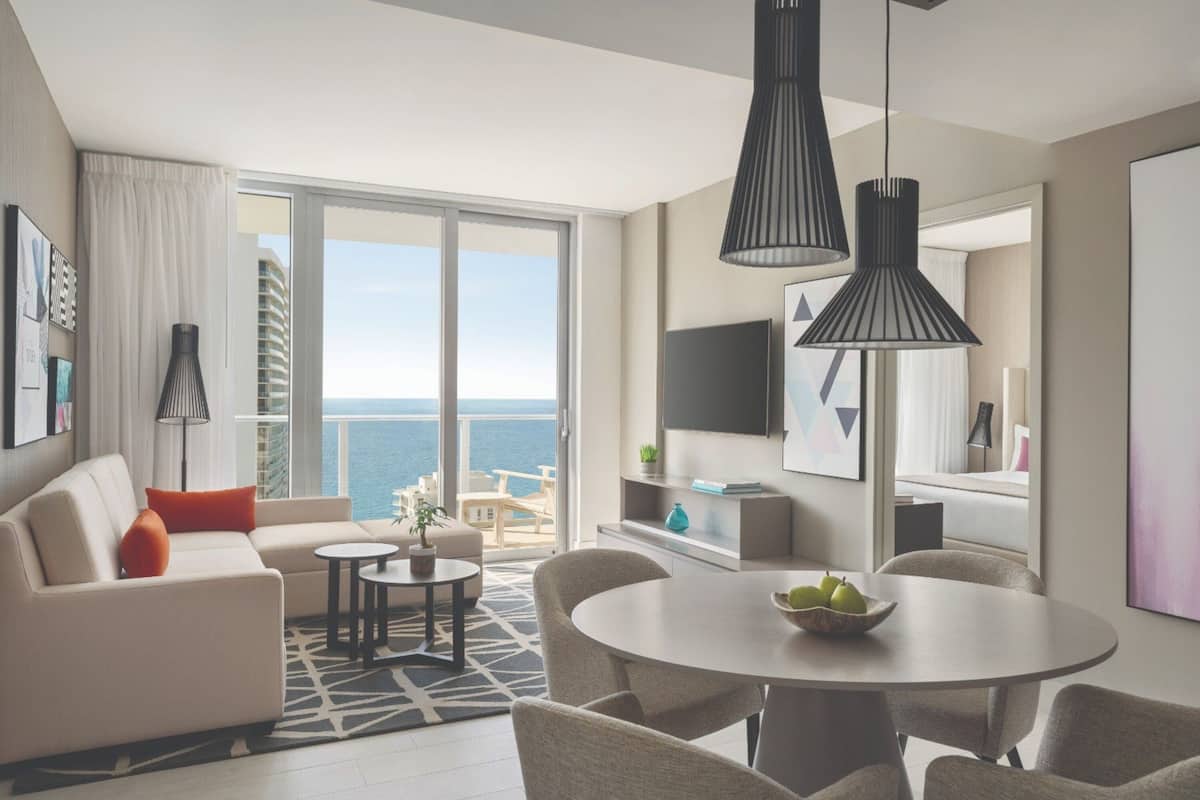 Uma sala de estar moderna com sofá, mesa de jantar e cadeiras, uma janela grande com vista para o mar, e uma varanda com mobiliário exterior.