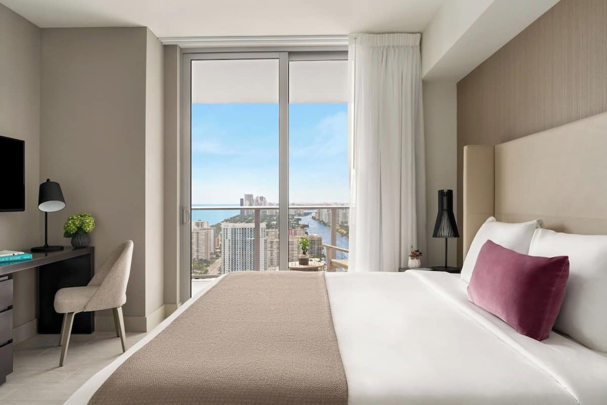 Quarto de hotel com uma cama grande, uma escrivaninha, uma cadeira, uma televisão e vista para a paisagem urbana.