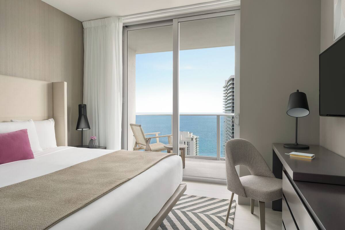 Quarto de hotel com cama, escrivaninha, poltrona, luminária e vista para o oceano e edifícios.