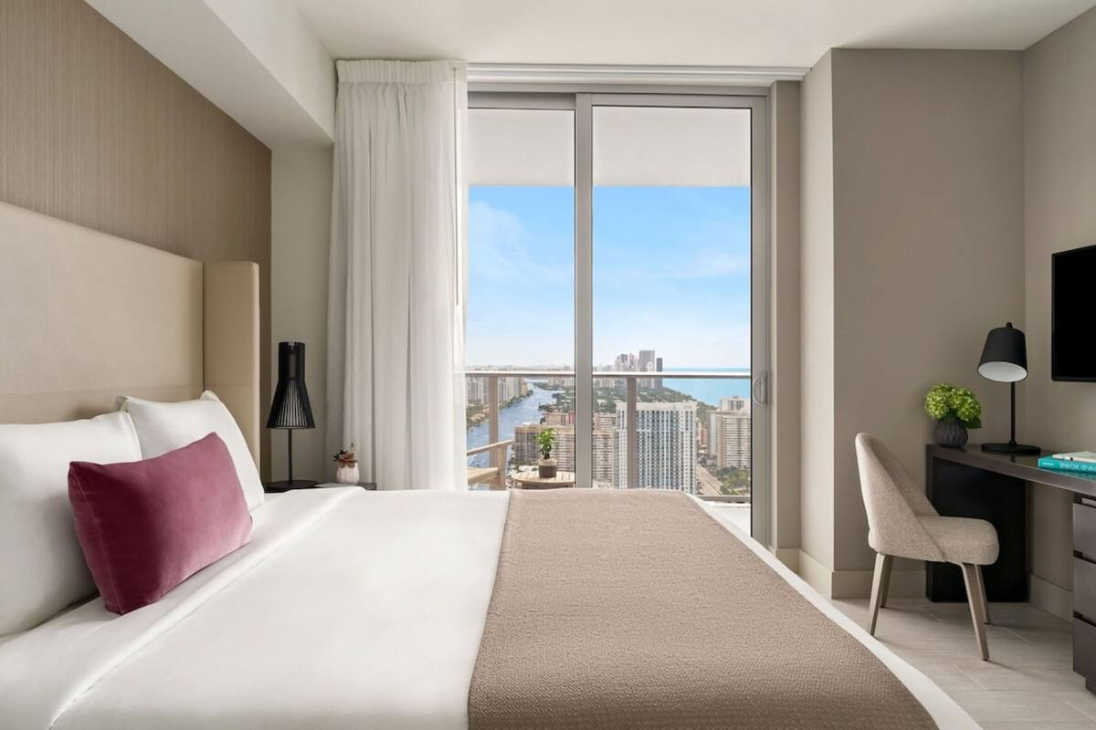 Quarto de hotel moderno com uma cama grande, uma escrivaninha, uma cadeira, uma televisão e vista para a cidade e para o mar.