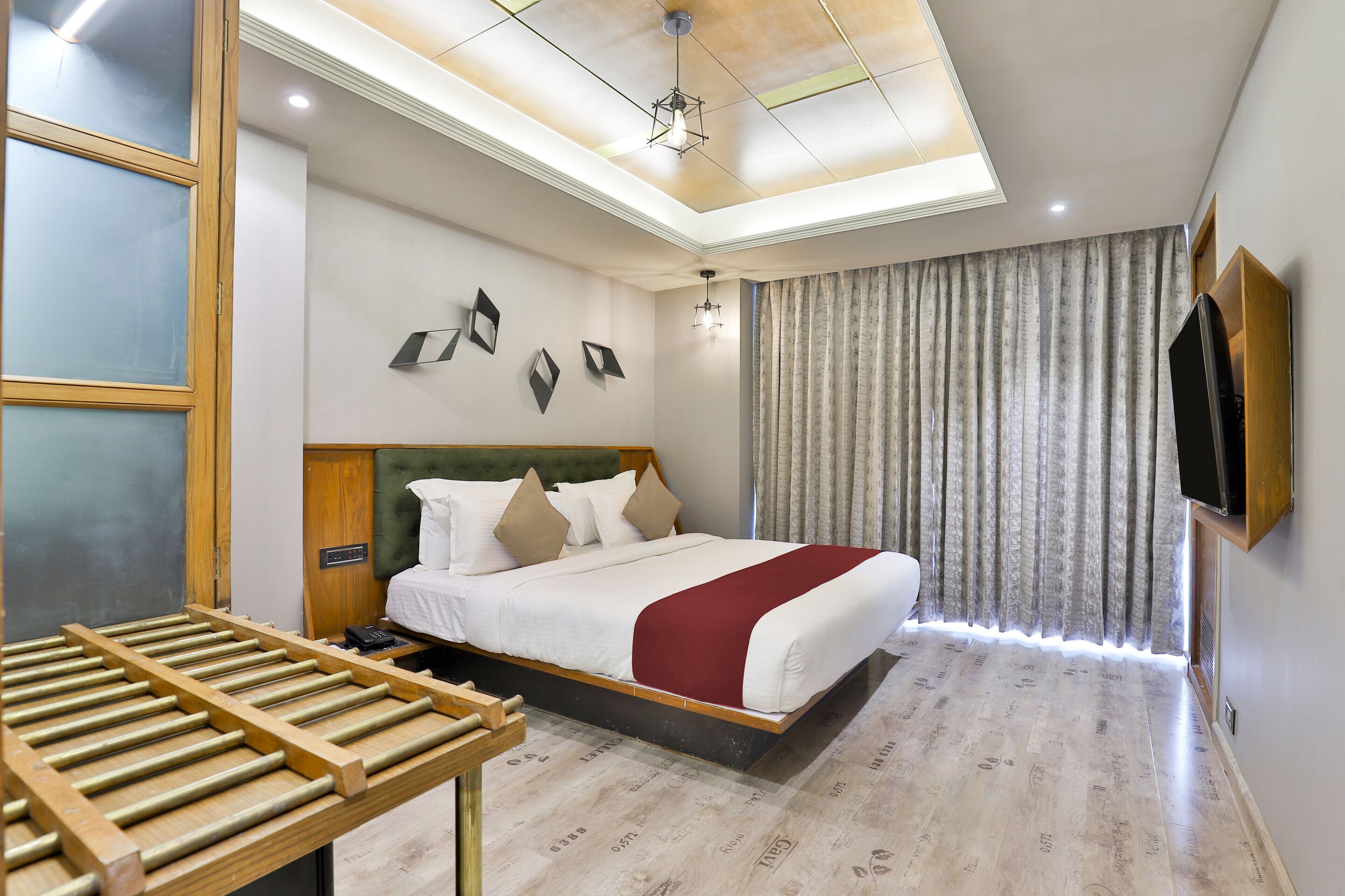 Quarto de hotel com uma cama grande, uma mesa de cabeceira de madeira, uma luminária no teto e uma televisão fixada na parede.