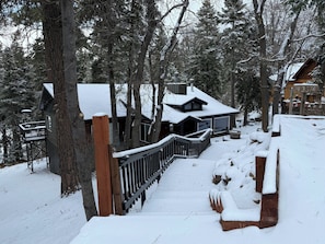 Uma cabana com deck de madeira e telhado coberto de neve, rodeada por árvores.