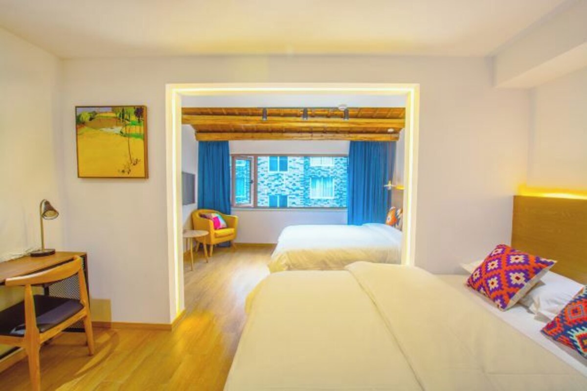 Quarto de hotel com duas camas, uma escrivaninha, uma cadeira e vista para uma paisagem montanhosa.