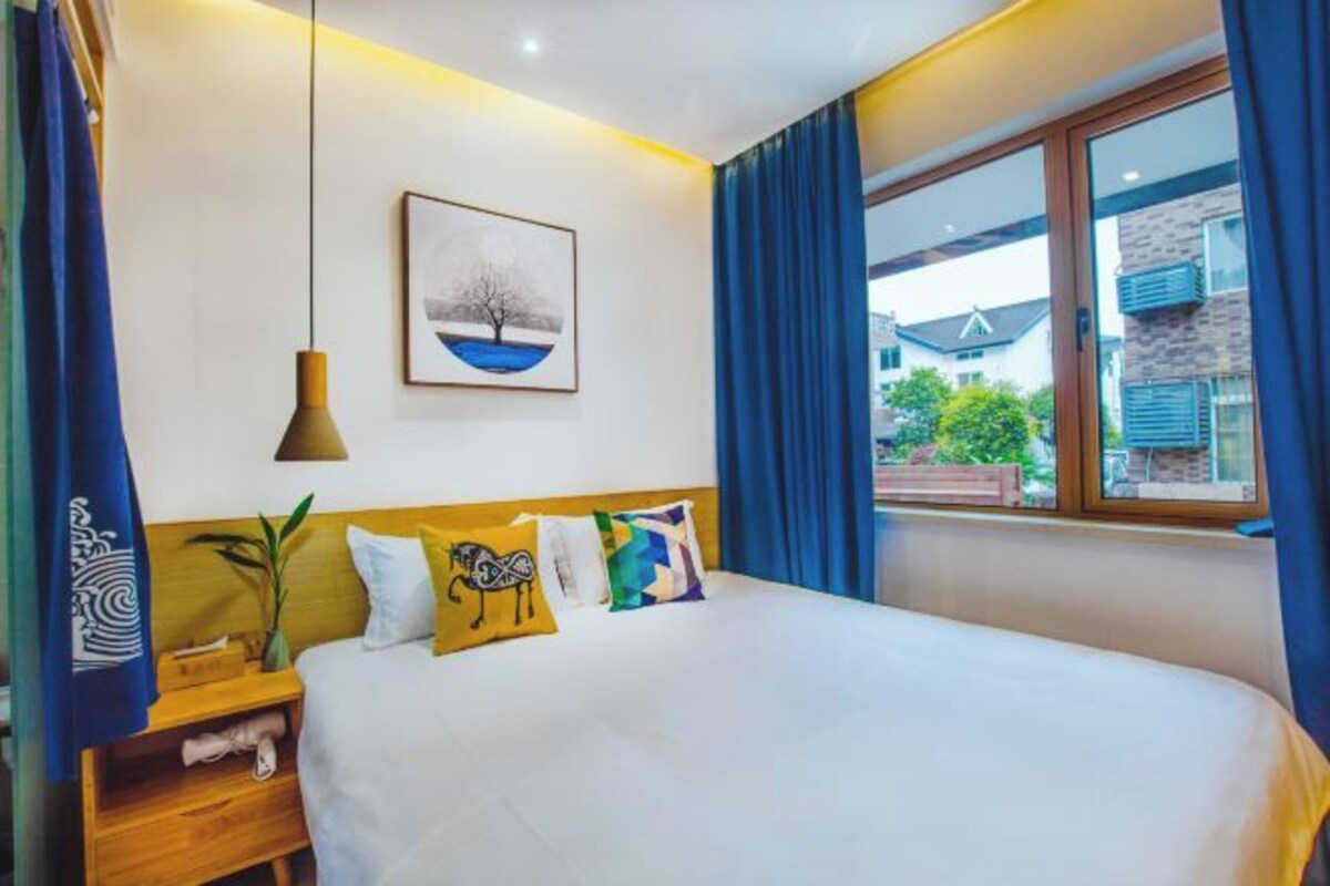 Quarto de hotel moderno com uma cama grande, mesa de cabeceira, luminária suspensa e janela com cortinas.
