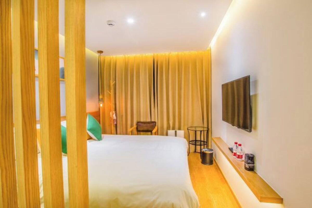 Quarto de hotel com uma cama, uma mesinha, uma cadeira, uma televisão na parede e uma cortina.