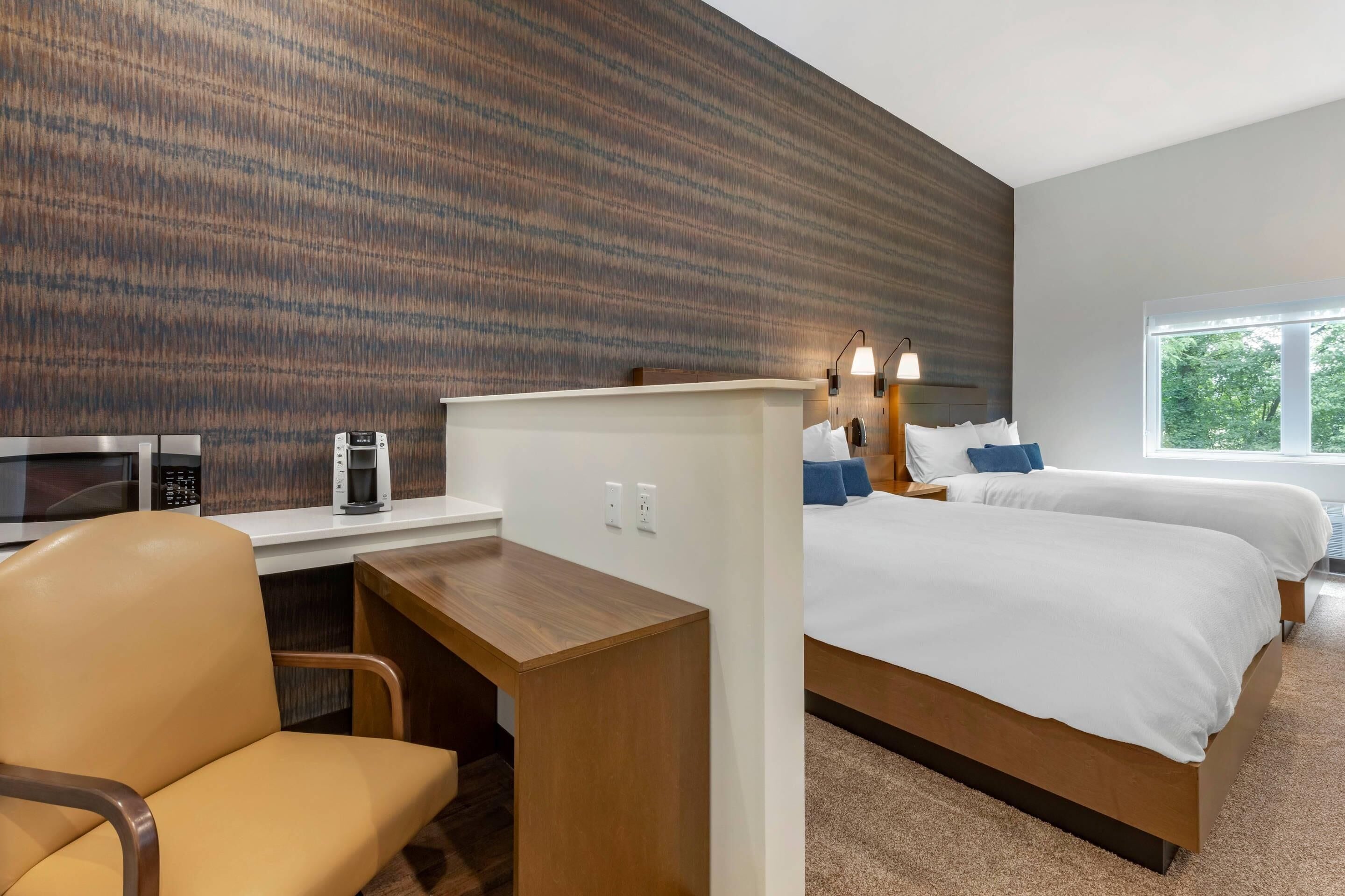 Quarto de hotel com secretária de madeira, uma cadeira, cama com roupa de cama branca, mesa de cabeceira com abajur e janela com vista para árvores.