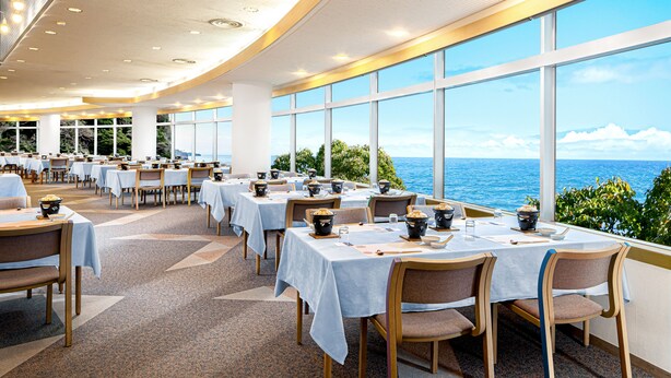 Restaurante com mesas preparadas para refeições, grandes janelas com vista para o mar e um design moderno no teto.
