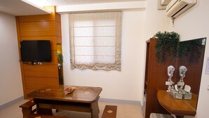 Quarto com mesa de madeira, bancos, televisão fixada na parede, janela com cortina estampada e espelho com planta na parede.