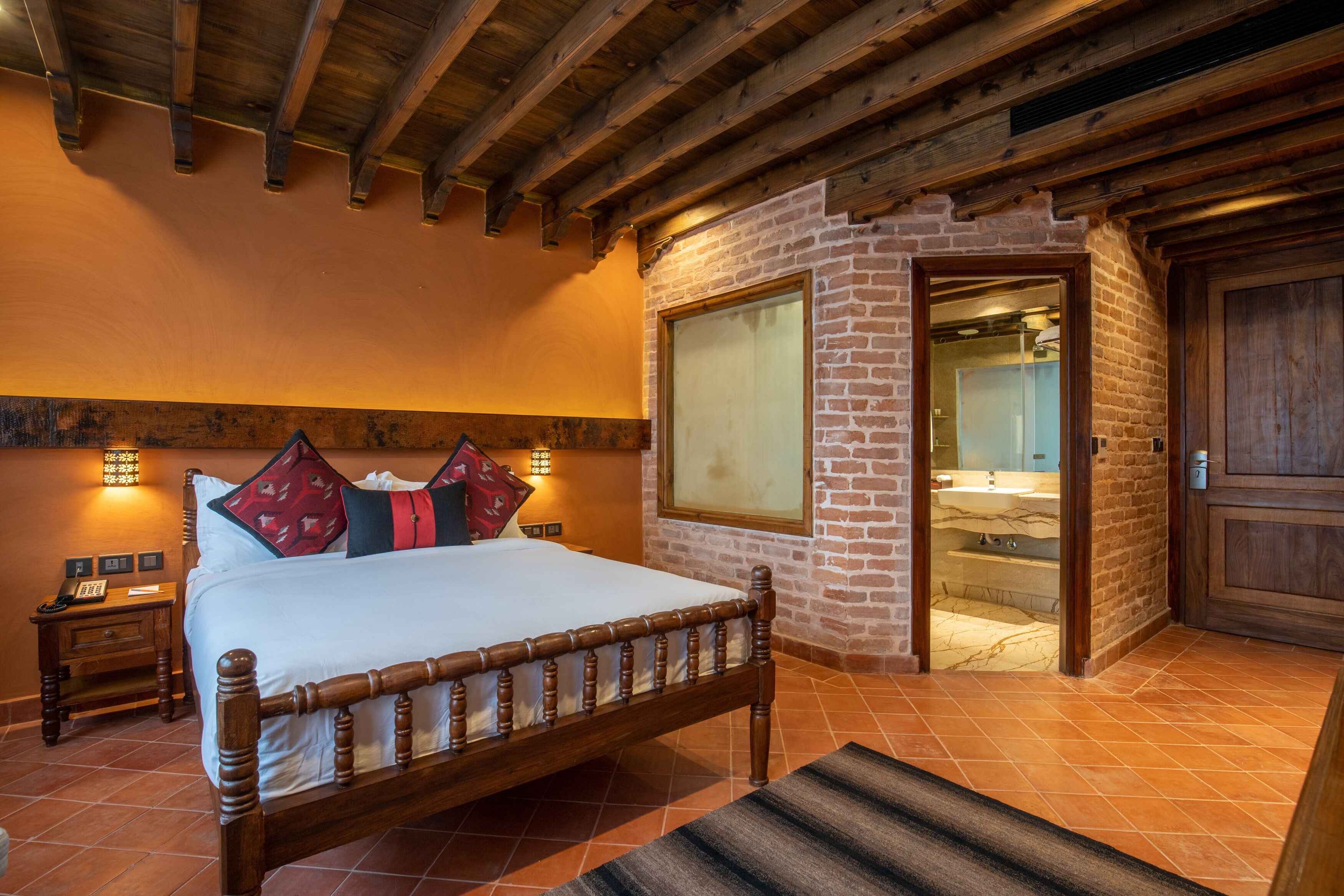 Uma cama de madeira com cabeceira, um criado-mudo com um telefone, e um banheiro visível por uma porta aberta.