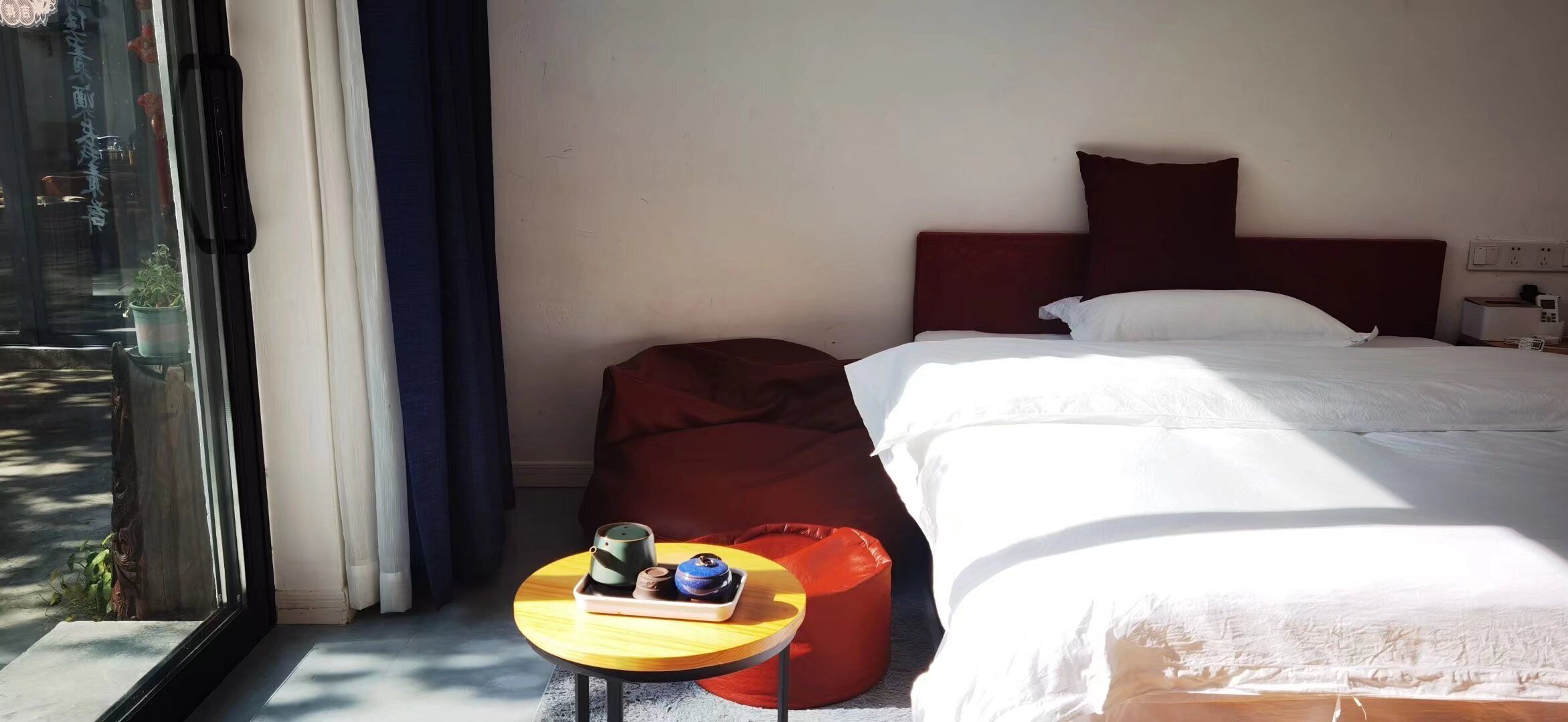 Quarto de hotel com uma cama, uma pequena mesa com uma bandeja de objetos, uma cadeira vermelha e uma janela com cortinas.