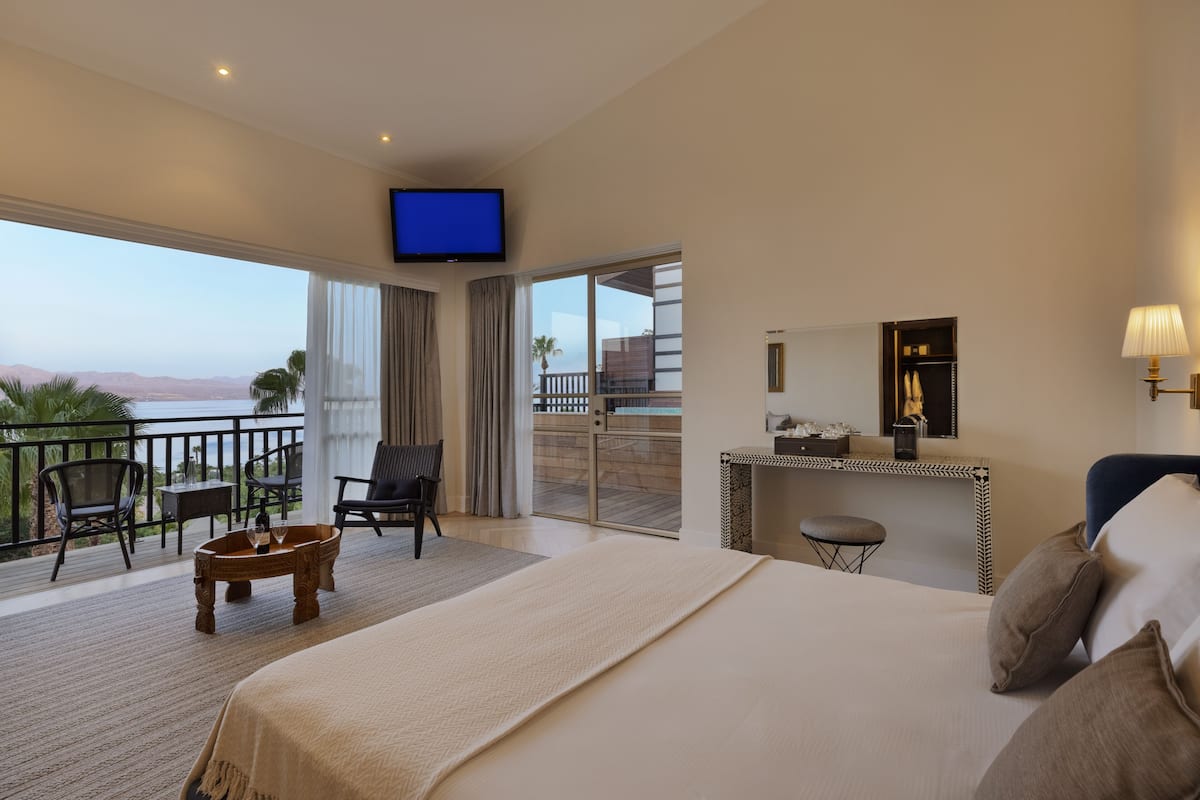 Quarto de hotel com uma cama grande, uma escrivaninha, televisão e varanda com cadeiras e mesa.