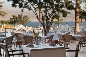 Um restaurante à beira-mar com mesas e cadeiras, uma garrafa de champanhe, e vista para o mar e montanhas.