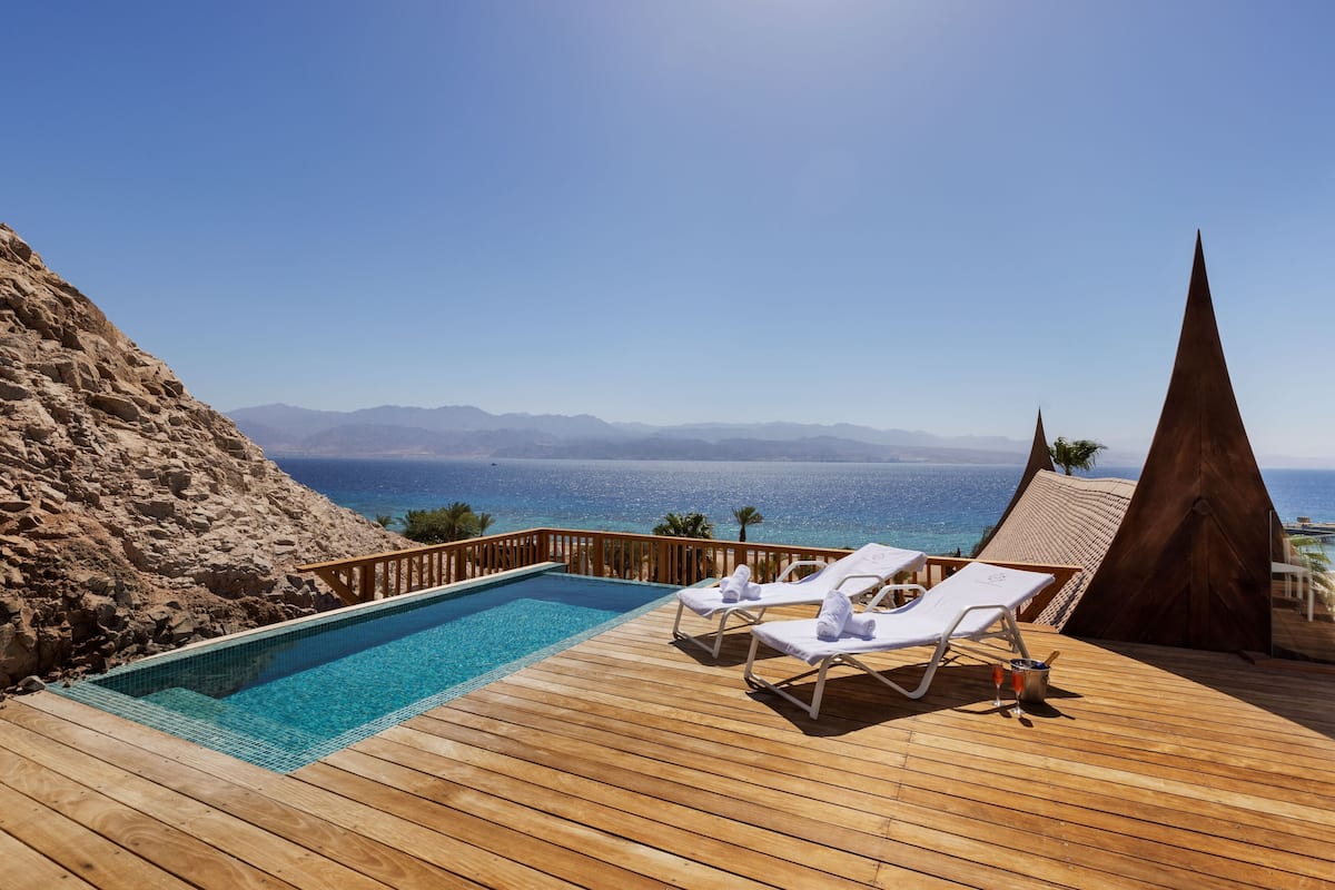 Um deck de madeira com piscina, duas espreguiçadeiras e vista para o mar e montanhas.