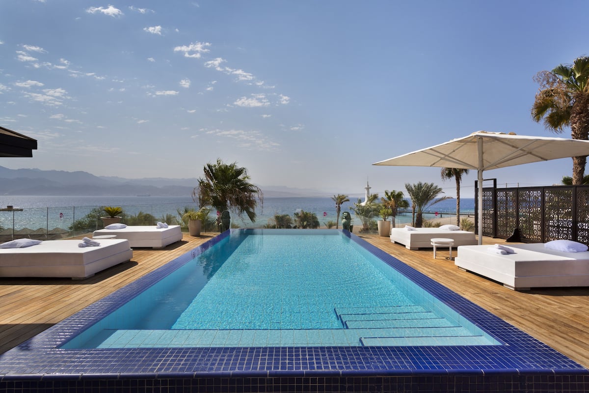 Piscina retangular com água azul cristalina, rodeada por um deck de madeira e espreguiçadeiras brancas.