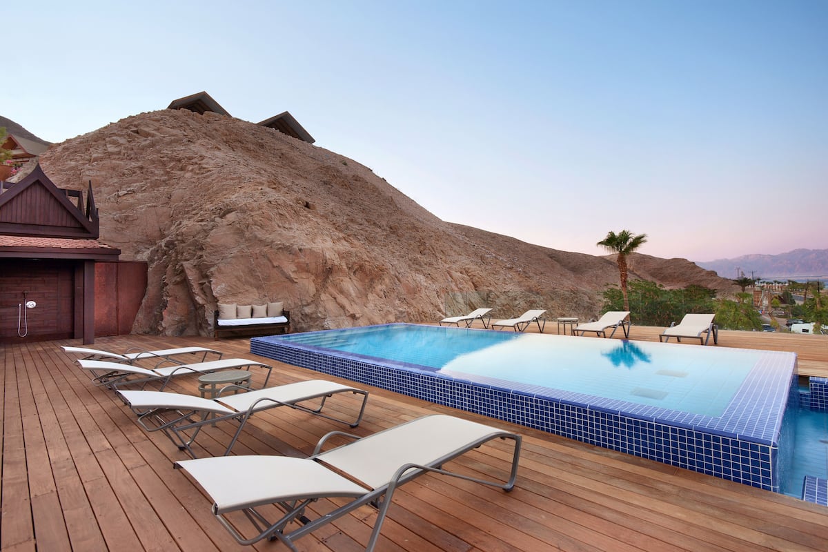 Área de piscina com espreguiçadeiras, deck de madeira e montanhas ao fundo.