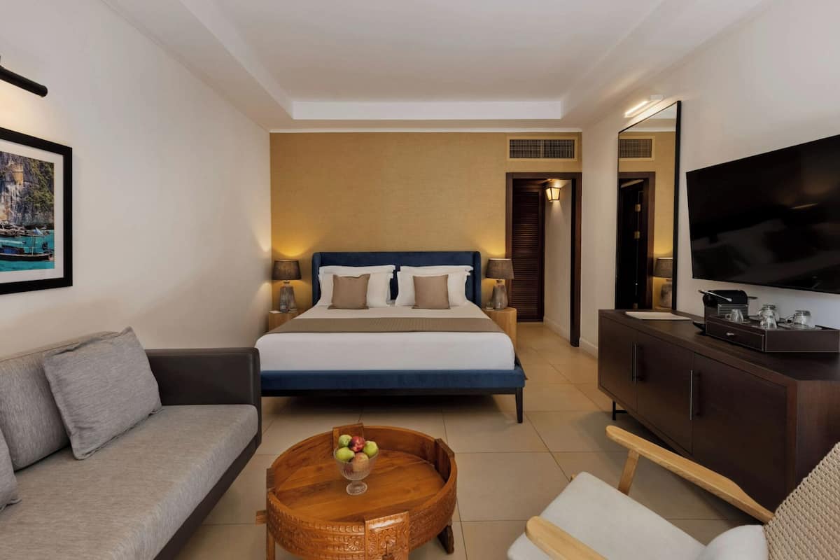 Quarto de hotel com cama, televisão, sofá, mesa de centro com frutas e um quadro na parede.