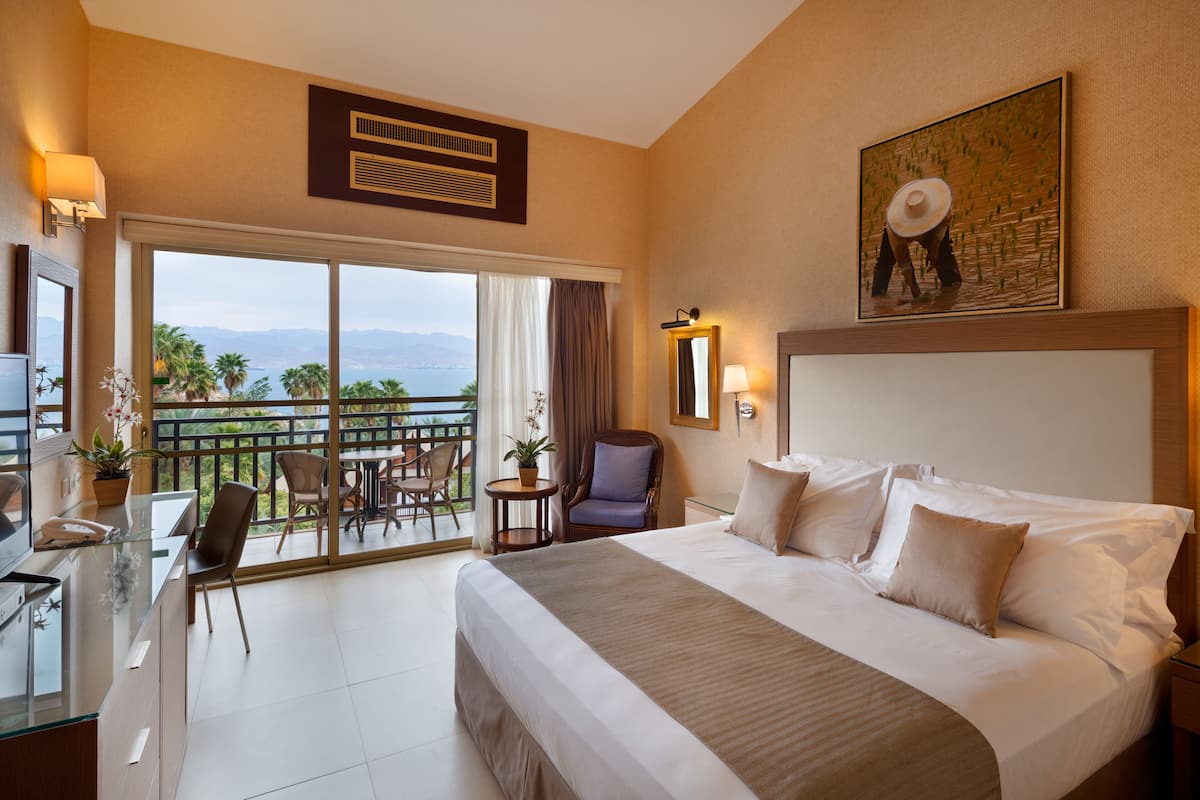Quarto de hotel com uma cama grande, uma mesa com cadeira, uma pequena mesa com uma planta, uma poltrona e uma varanda com vista para as montanhas.
