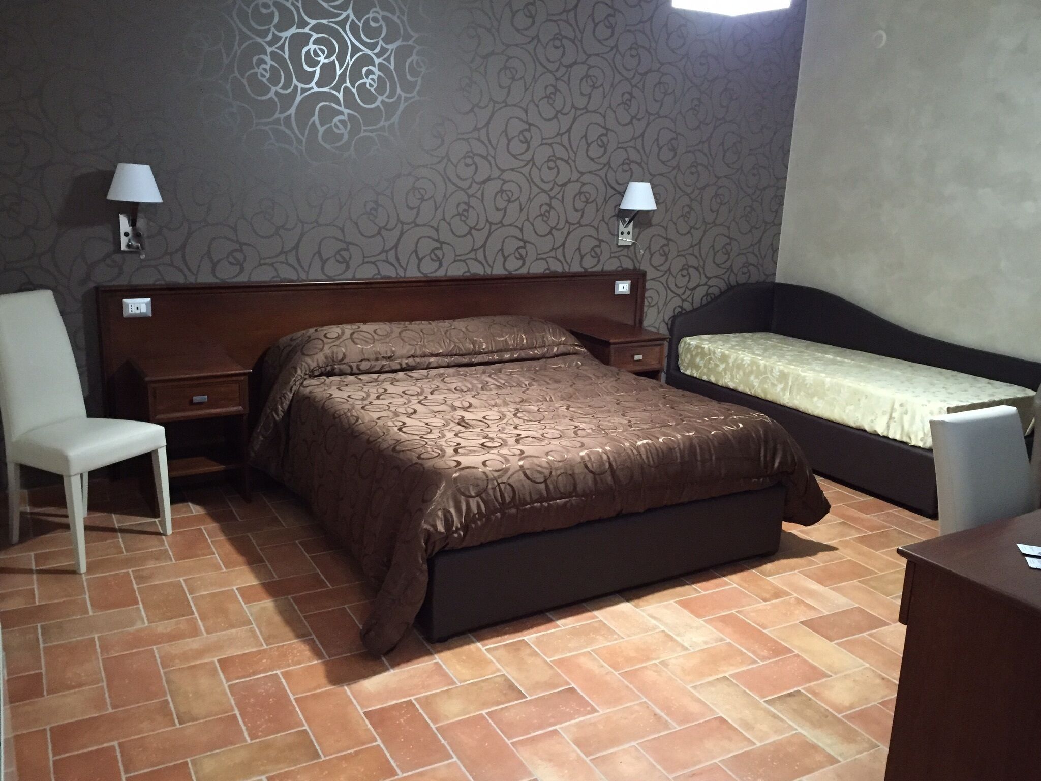 Quarto com uma cama grande, duas mesas de cabeceira, uma cadeira e papel de parede estampado.