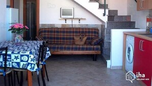 Uma sala de estar com sofá, mesa de jantar e cadeiras, escada, máquina de lavar e uma pequena TV na parede.