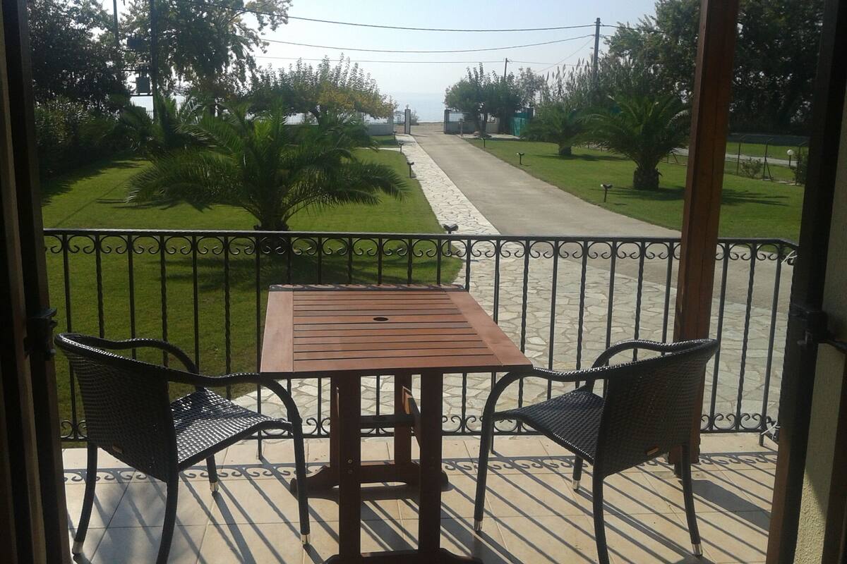 Varanda com mesa e duas cadeiras, com vista para um caminho pavimentado e vegetação.