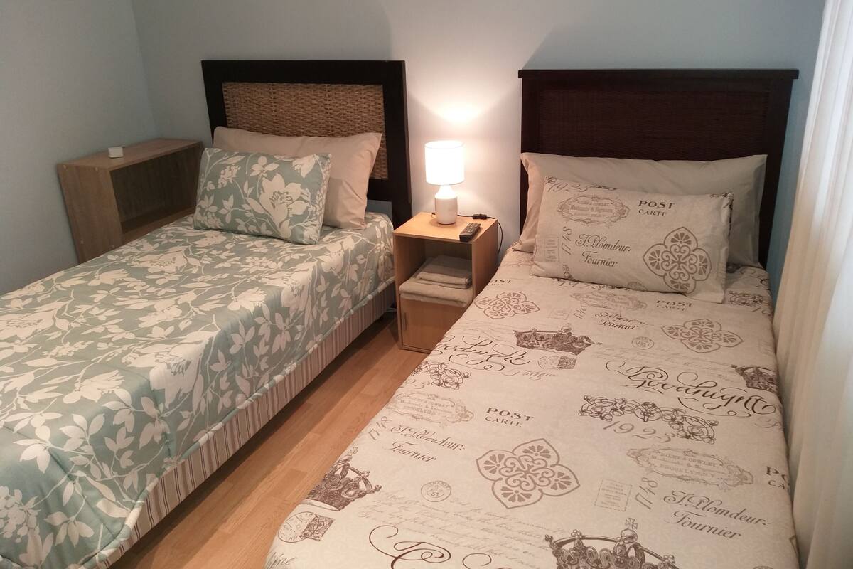 Quarto duplo com duas camas, cada uma com roupa de cama florida e cabeceira. Há uma mesa de cabeceira com abajur entre as camas.