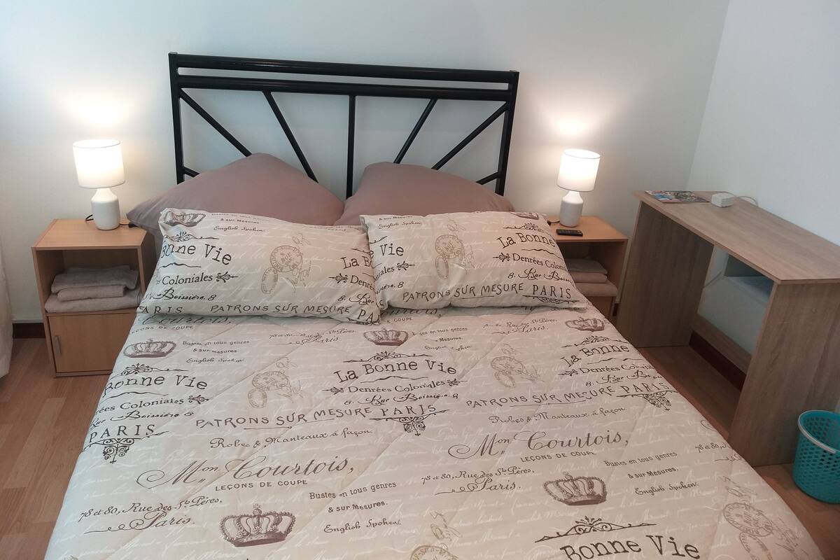 Cama bem arrumada com cabeceira, duas luminárias de apoio e mesa de cabeceira de madeira com luminária.
