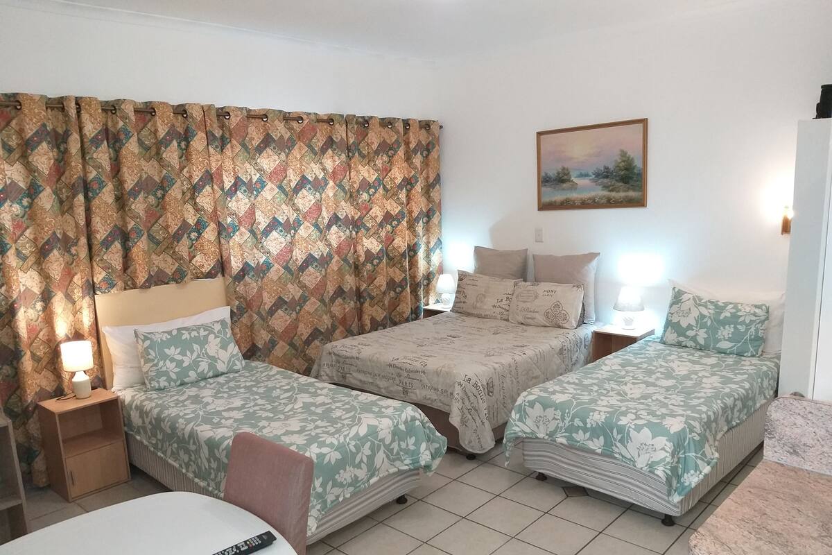 Quarto de hotel com duas camas de solteiro, uma mesinha, uma cadeira, um abajur e um quadro na parede.