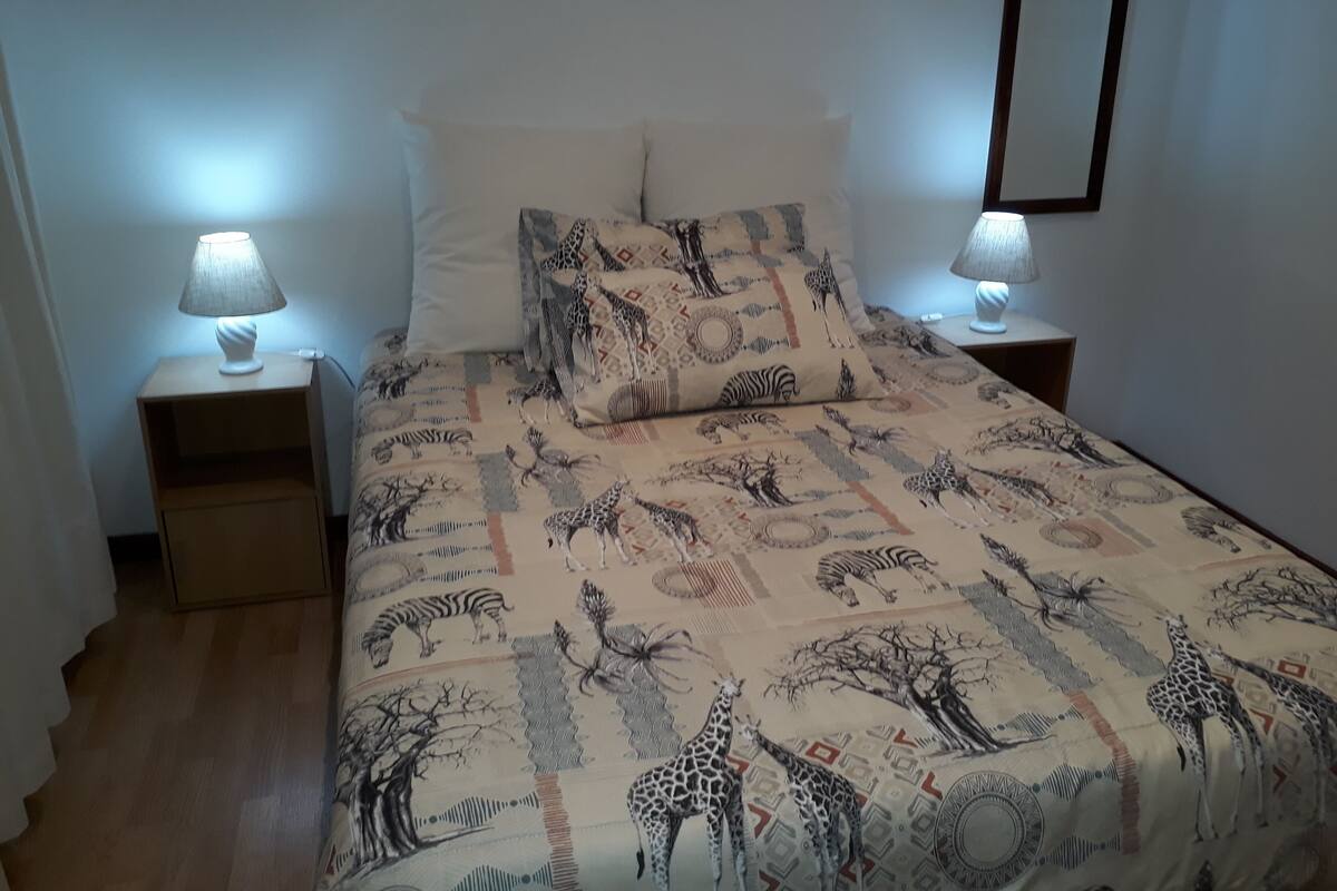 Quarto com colcha estampada, duas luminárias de cabeceira e uma mesa de apoio.