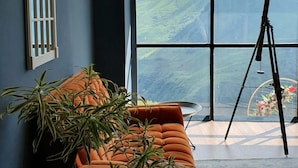 Quarto com uma grande janela com vista para as montanhas, um tripé com uma câmera e uma planta em vaso em um sofá moderno.