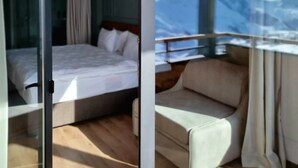 Quarto de hotel moderno com uma cama grande, um sofá e vista para montanhas através de uma janela ampla.