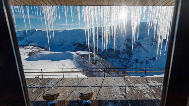 Um deck de madeira com duas cadeiras de vime, com vista para uma paisagem montanhosa coberta de neve e pingentes de gelo no teto.