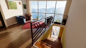 Quarto espaçoso com piso de madeira, tapete vermelho, uma cadeira azul e uma grande janela com vista para montanhas.
