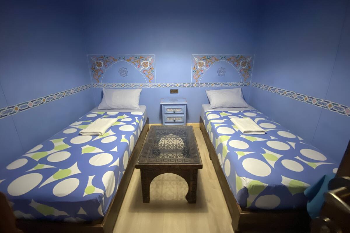 Quarto com duas camas de solteiro, cada uma com colcha estampada, uma pequena mesa de madeira e azulejos decorativos na parede.