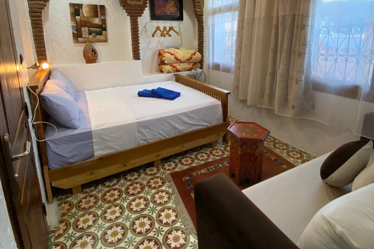 Quarto com cama de madeira, uma mesinha, tapete decorativo, janela com cortinas e luminária suspensa.