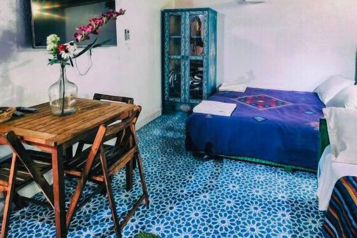 Quarto com mesa de jantar de madeira, cadeiras, cama com colcha azul e roxa, e armário com portas de vidro.